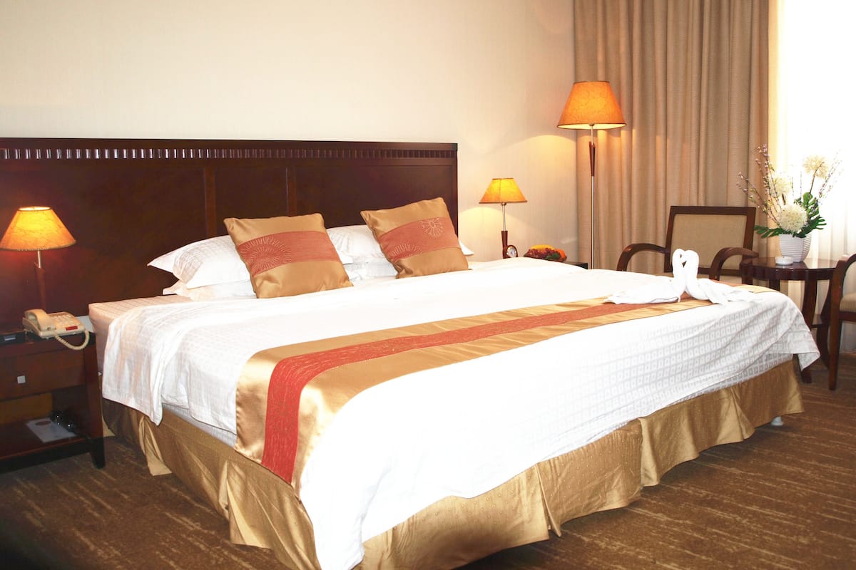 Quarto de hotel com uma cama grande, duas luminárias laterais, mesa de cabeceira com telefone, cabeceira de madeira, escrivaninha com cadeira e um vaso com flores.