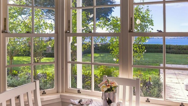 Uma sala de jantar com mesa posta para quatro pessoas, grandes janelas com vista para um jardim e o mar.