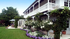 Uma casa de dois andares com varanda, rodeada por um gramado bem cuidado e canteiros de flores.