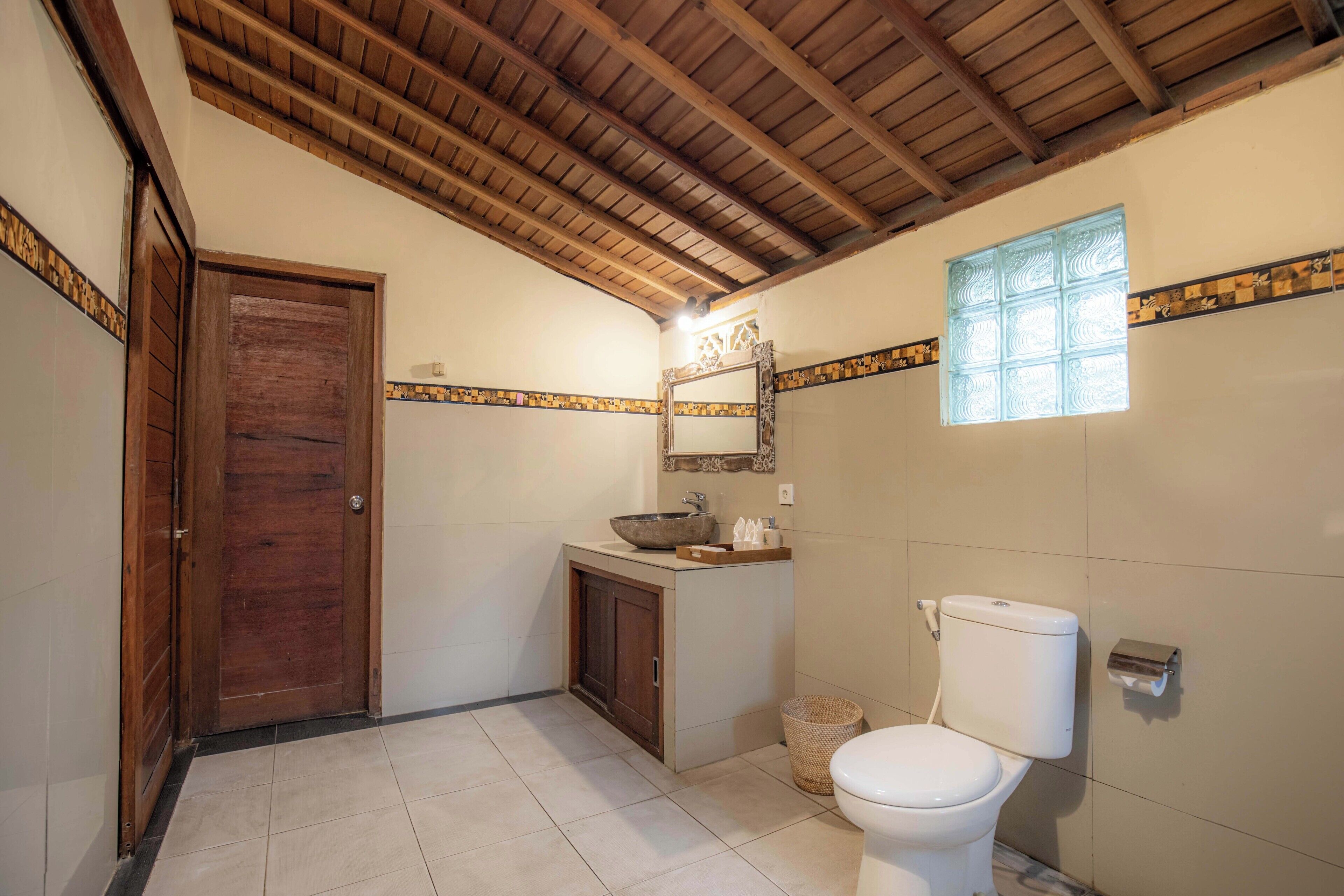 Um banheiro com porta de madeira, pia com espelho decorativo, vaso sanitário e janela com vidro estampado.