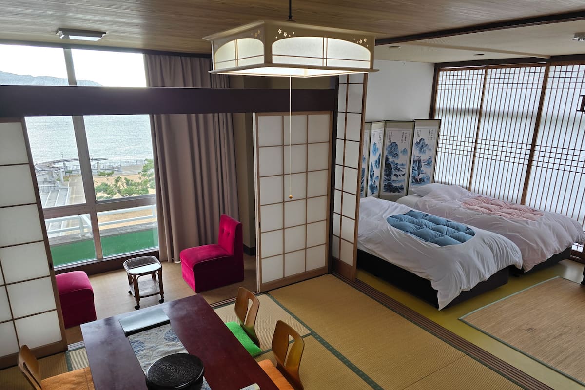 Quarto de hotel com uma grande janela, uma cama com edredom branco e rosa, uma mesa de jantar com um controle remoto e uma cadeira vermelha.