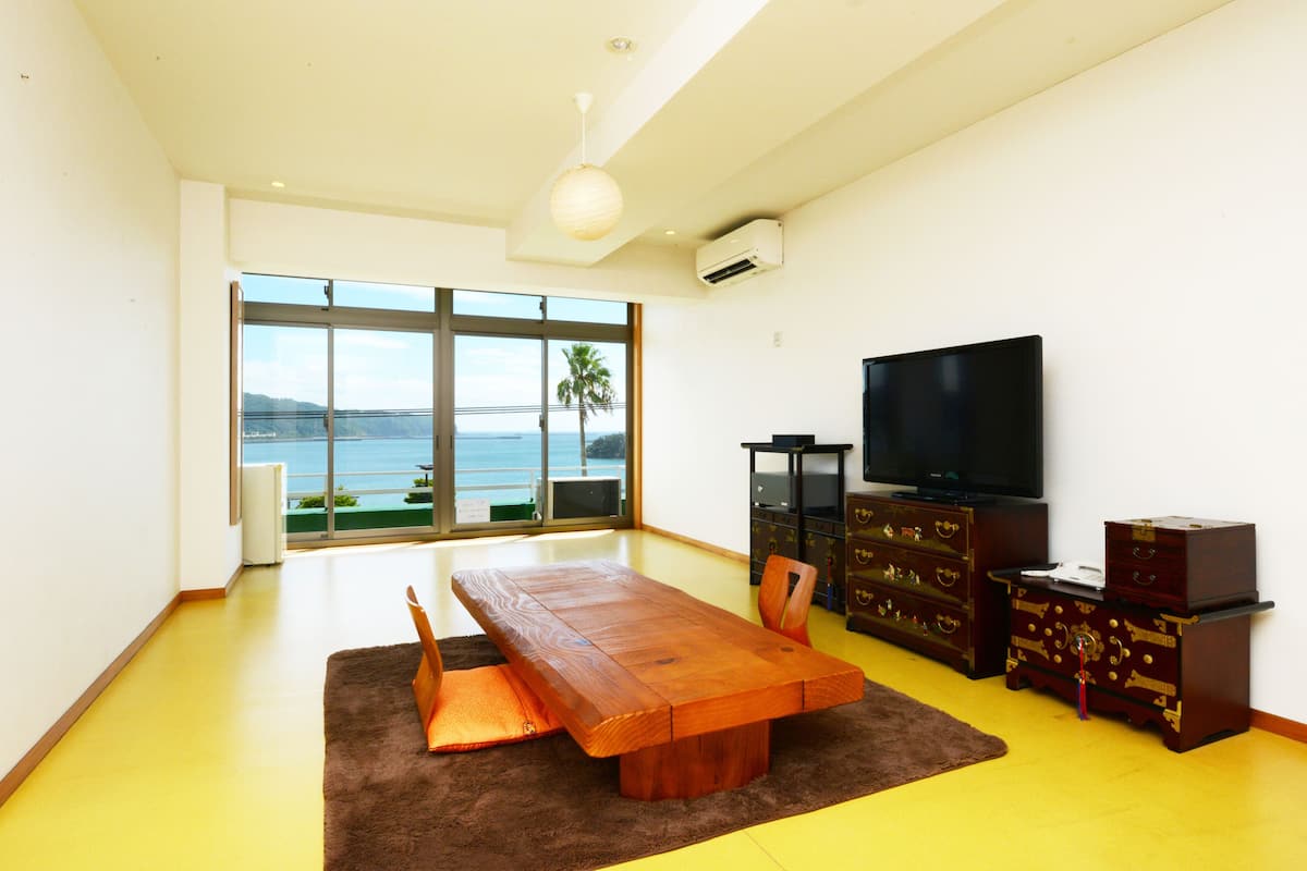 Sala de estar moderna com uma grande mesa de madeira, uma TV de tela plana e vista para o mar através de amplas janelas.