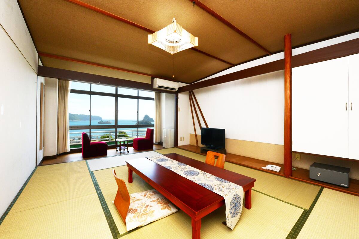 Um quarto japonês tradicional com tatames, uma mesa de madeira baixa, uma televisão e vista para o mar.
