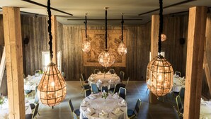Uma sala de jantar com mesas redondas preparadas para uma refeição, rodeada por cadeiras, com luminárias penduradas acima.
