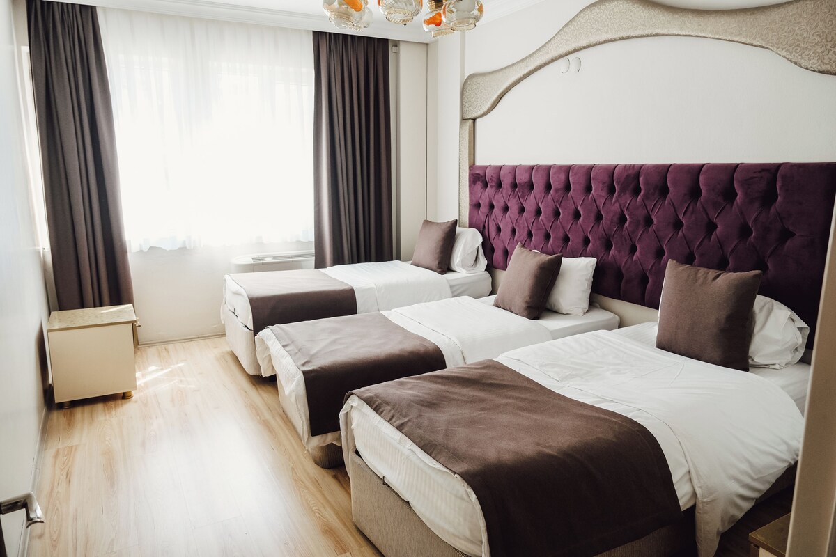 Quarto de hotel com duas camas, piso de madeira, uma janela grande com cortinas e cabeceira decorativa.