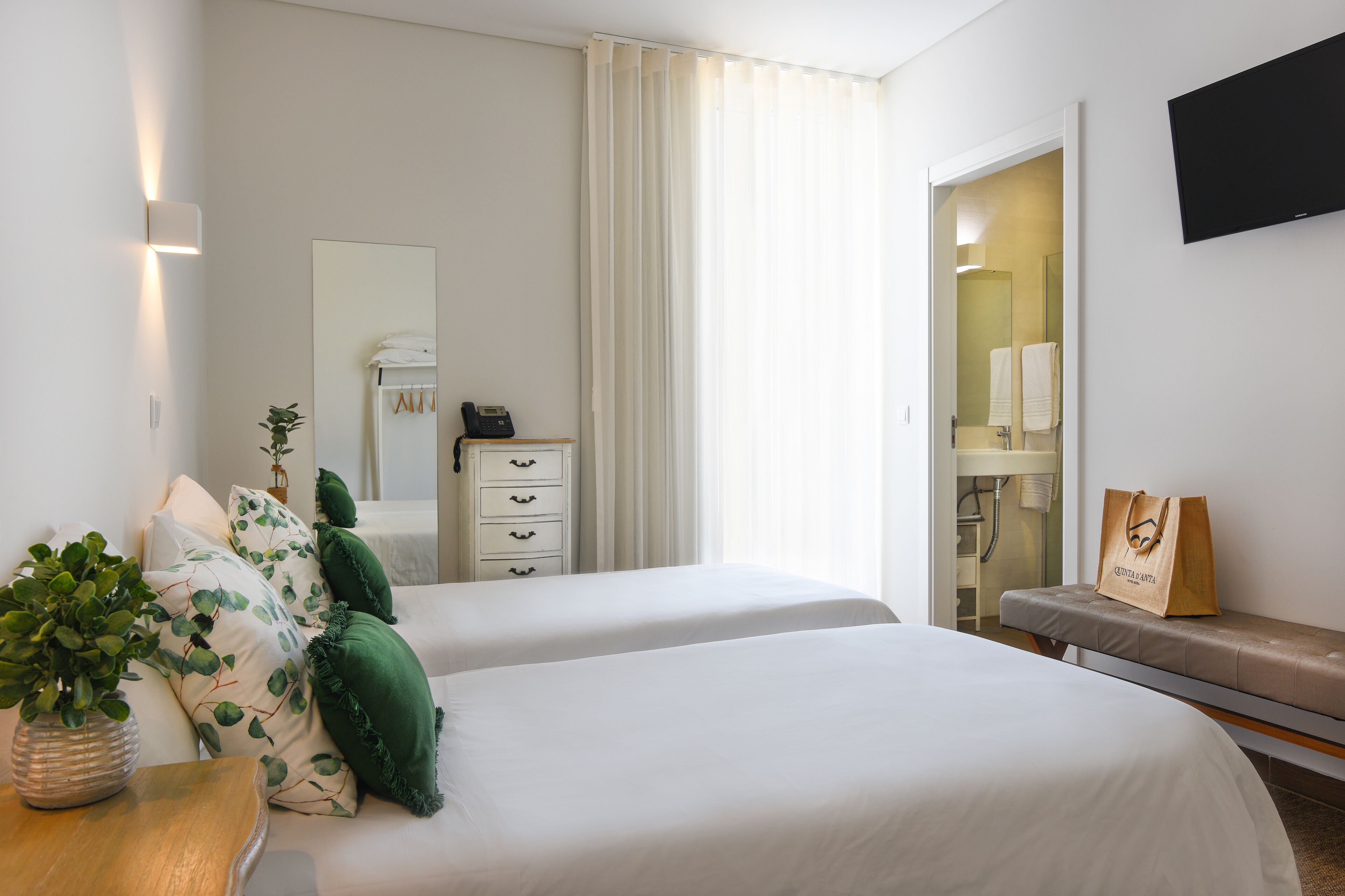 Quarto de hotel com duas camas, uma mesa de cabeceira com planta, um banco, um guarda-roupa, um espelho e uma televisão na parede.