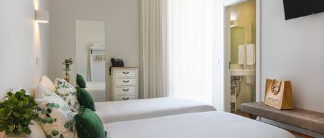 Quarto de hotel com duas camas, uma mesa de cabeceira com planta, um banco, um guarda-roupa, um espelho e uma televisão na parede.