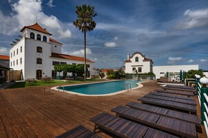 Um deck de madeira com piscina, espreguiçadeiras e dois edifícios brancos com telhados de telha vermelha.