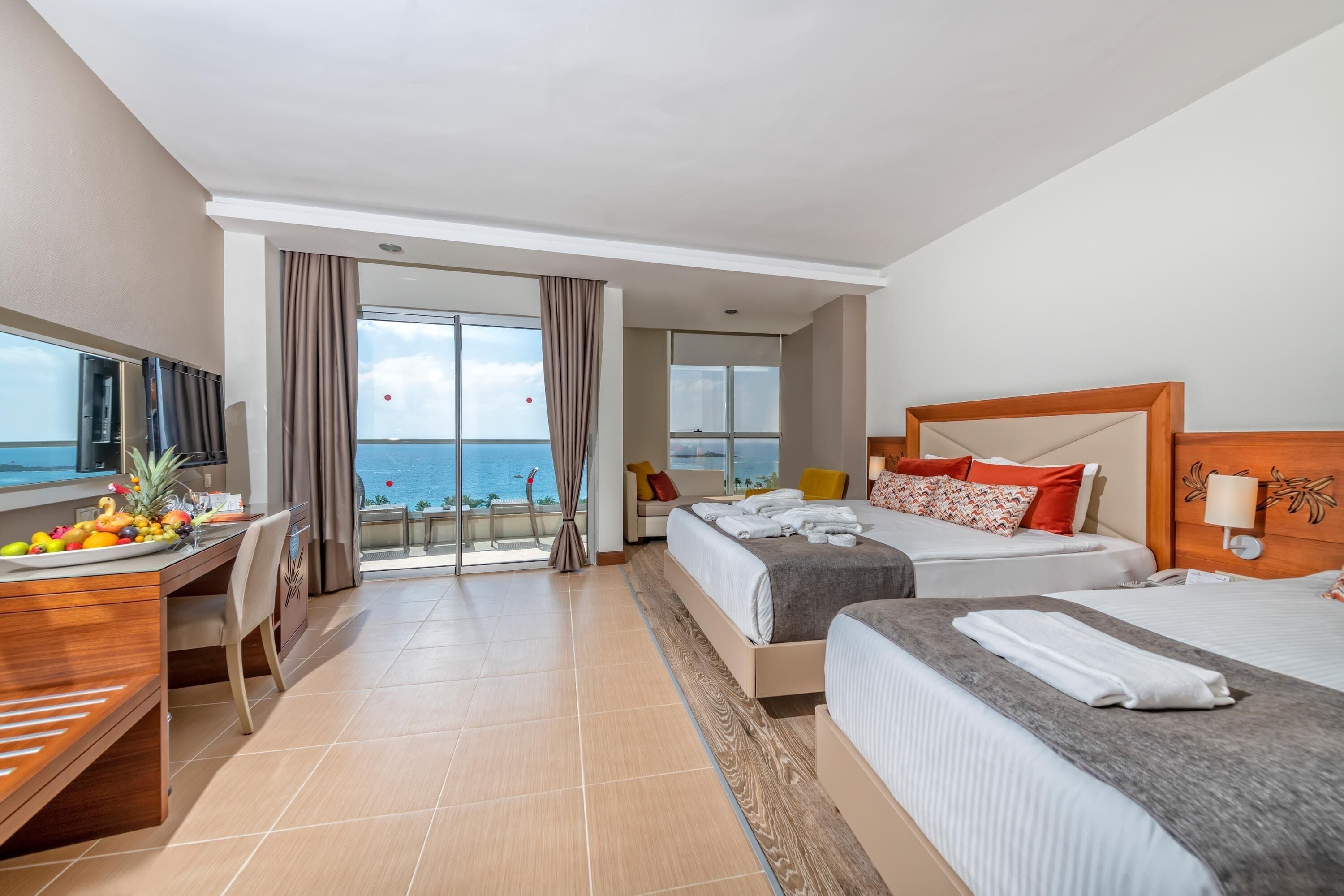 Quarto de hotel com varanda, mesa com frutas, duas camas e uma televisão.