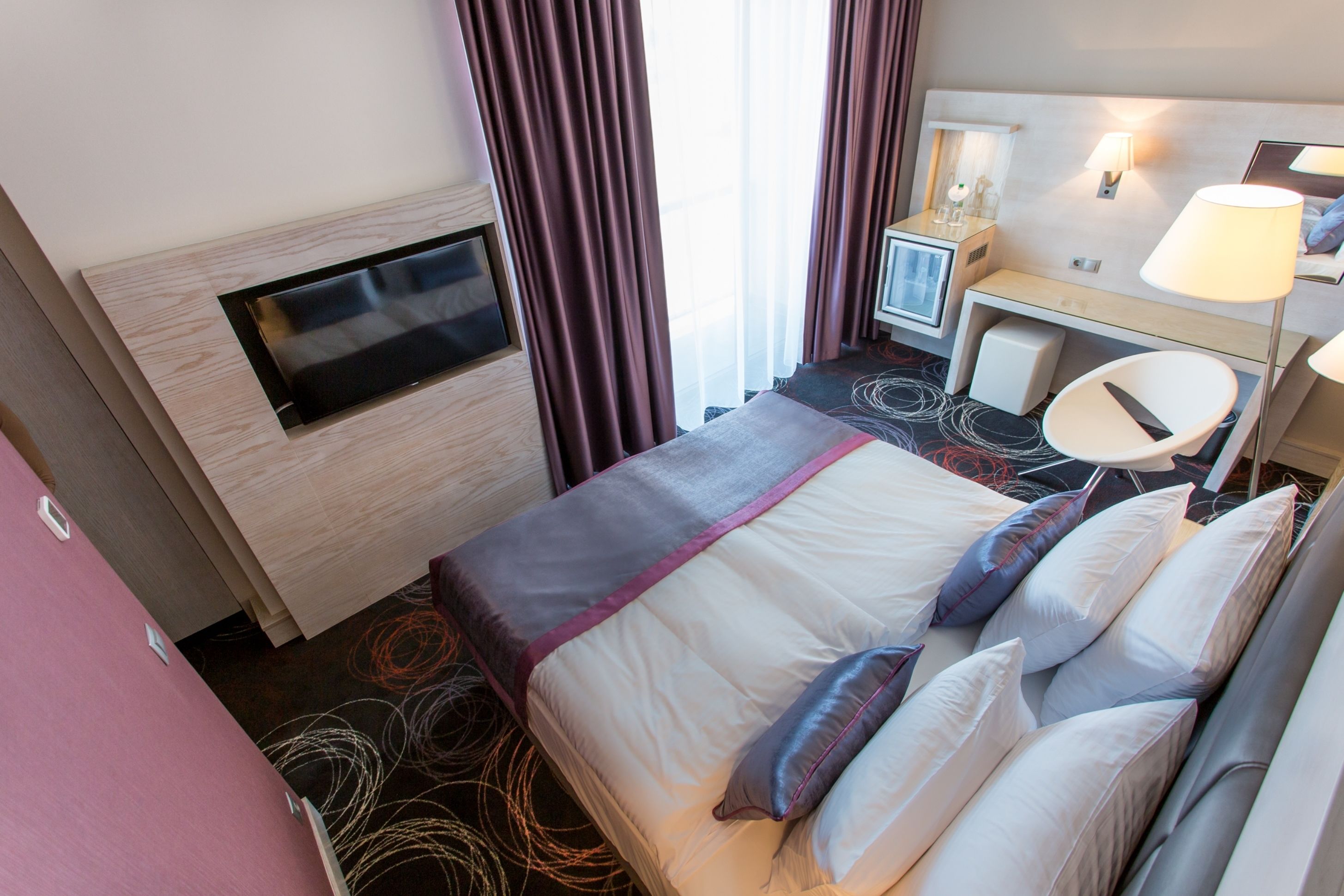 Quarto de hotel com uma cama, uma televisão, uma escrivaninha e uma luminária.