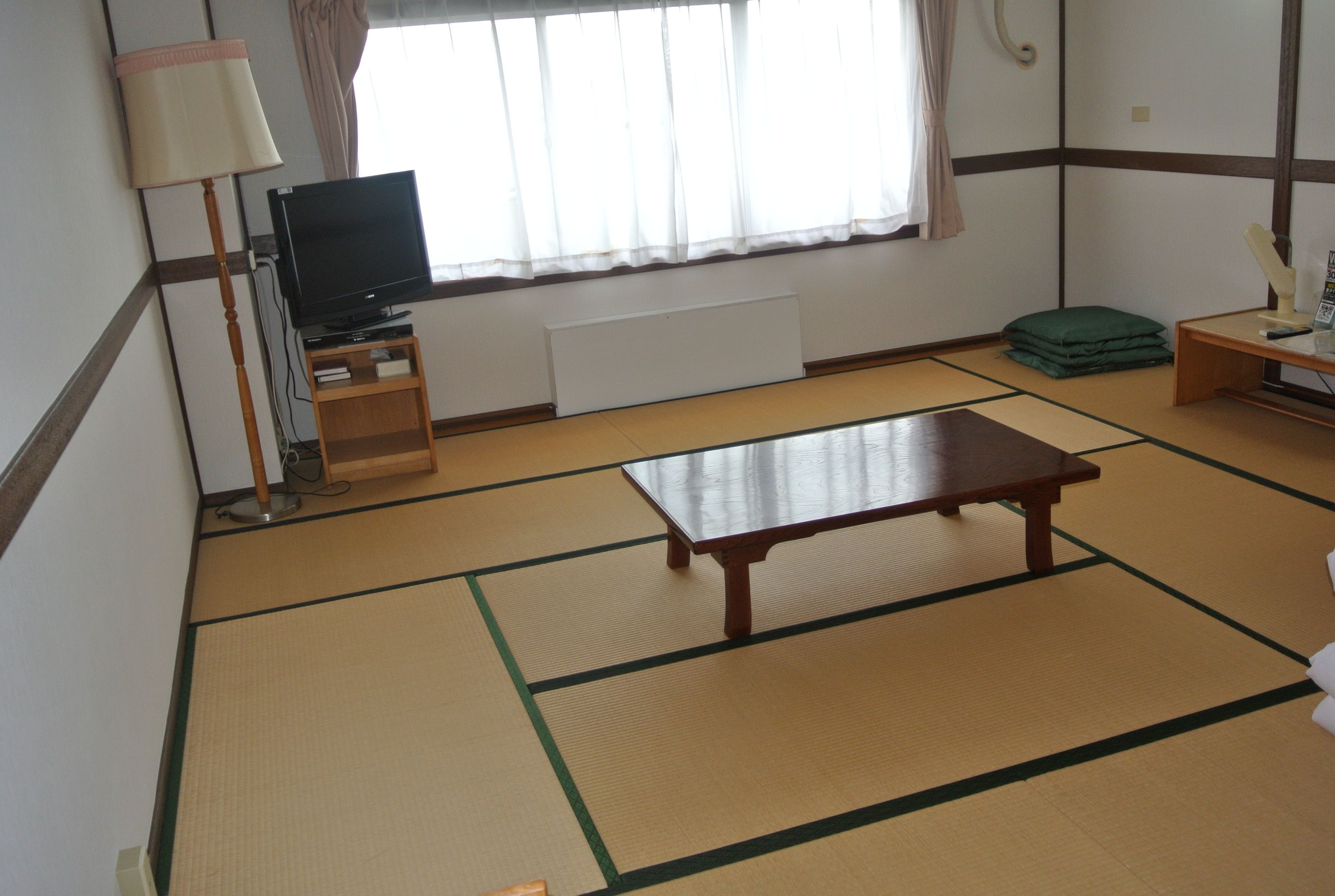 Quarto com piso de tatame, televisão em suporte, mesa e janela com cortinas.