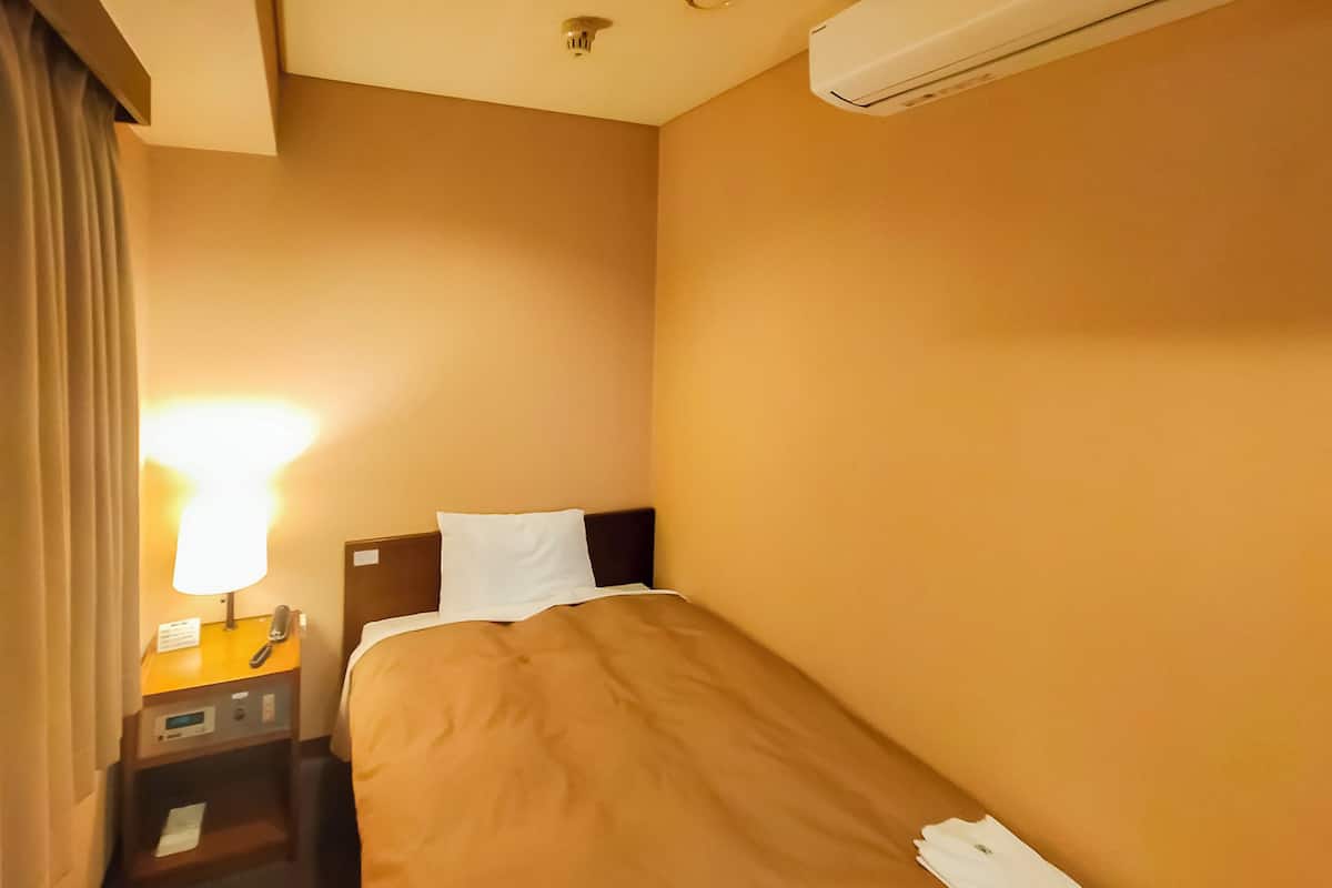 Quarto de hotel com uma cama de solteiro, mesa de cabeceira com abajur, telefone e ar condicionado fixado na parede.