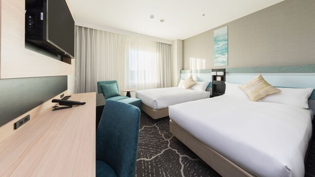 Quarto de hotel com duas camas, uma escrivaninha, uma cadeira, uma televisão e uma janela com cortinas.