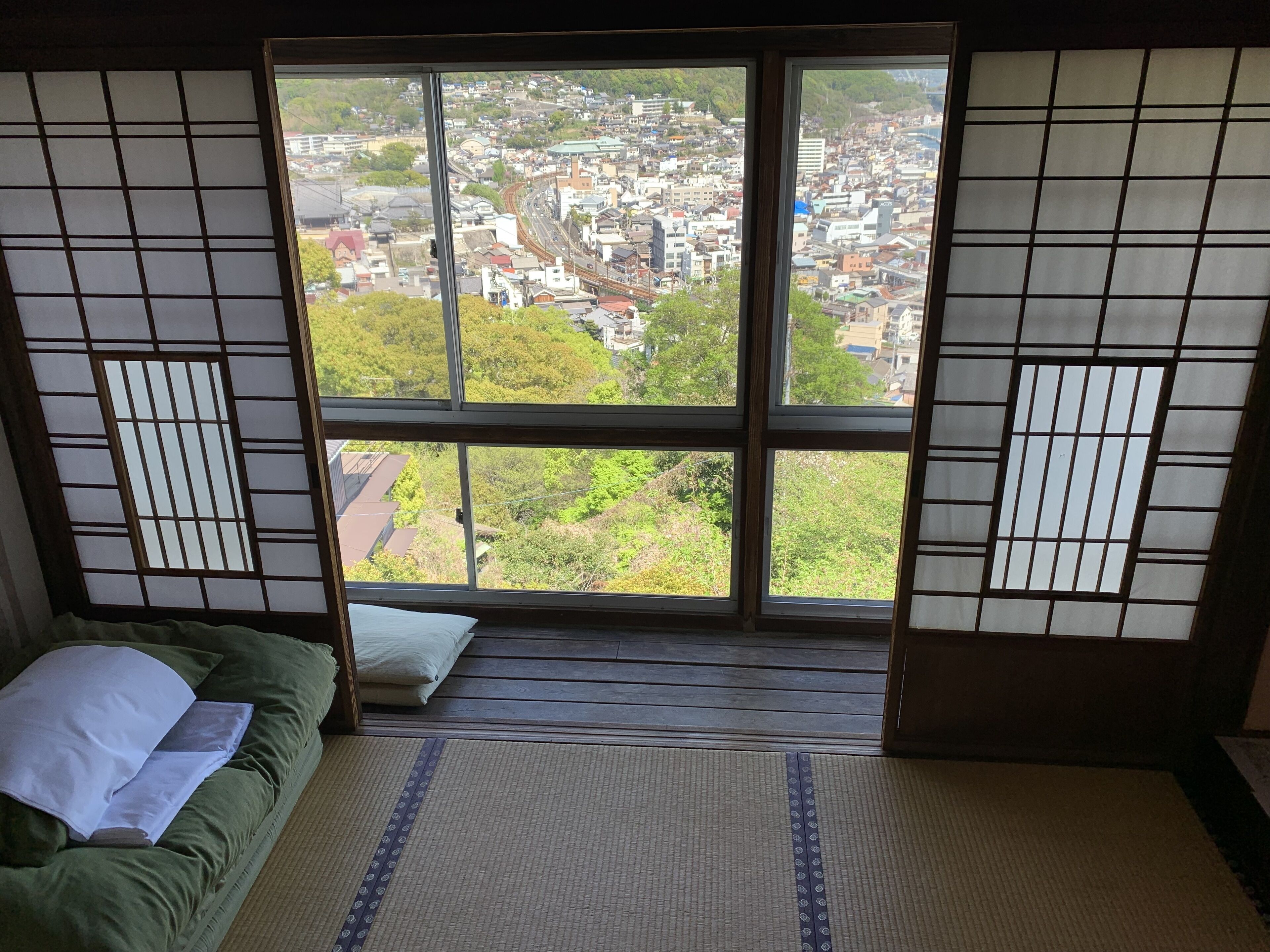 Um quarto japonês tradicional com piso de tatame, um sofá verde baixo com almofadas e uma grande janela com vista para a paisagem urbana.