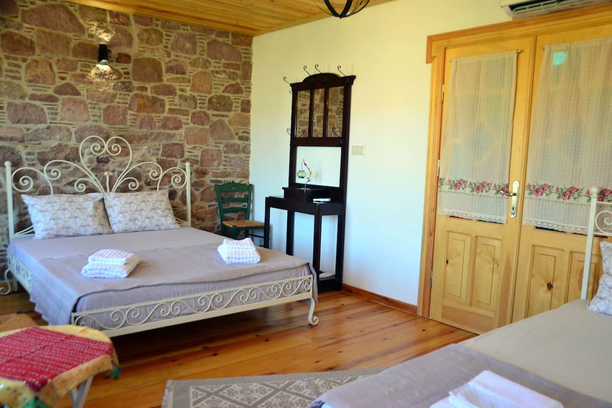 Um quarto com parede de pedra, porta de madeira com cortina floral, cama com estrutura de metal e uma pequena mesa com um espelho.