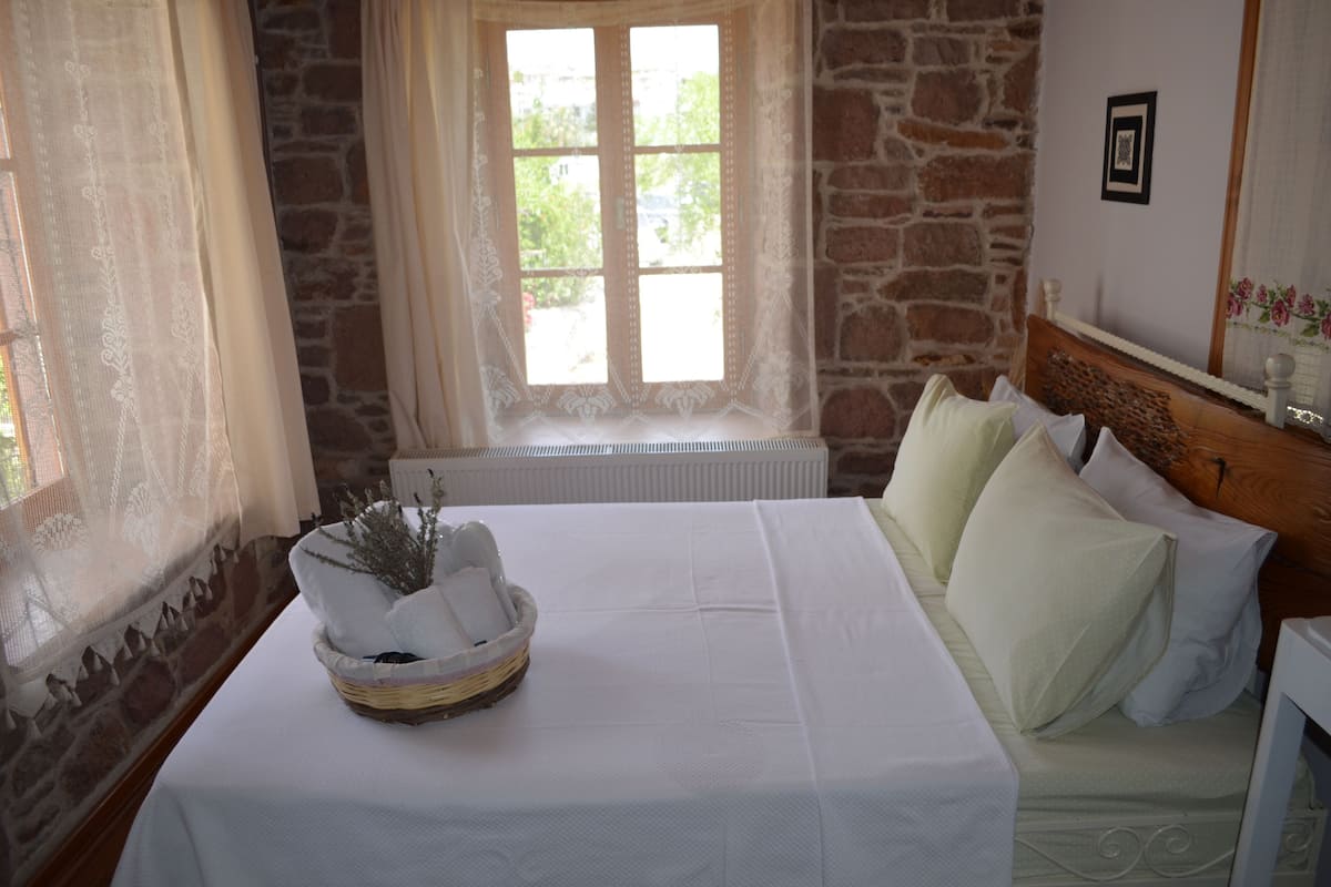 Quarto com parede de pedra, uma janela grande com cortinas, cama com roupa de cama branca e um cesto com uma planta sobre a cama.