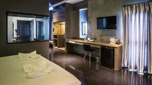 Quarto de hotel moderno com uma cama grande, uma escrivaninha, uma cadeira, televisão e banheiro visível através de uma porta aberta.