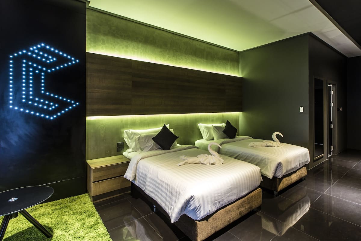 Quarto de hotel com duas camas, uma parede de destaque verde, um letreiro neon e uma mesa pequena.