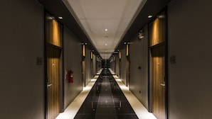 Corredor longo e estreito de hotel, com portas em ambos os lados, chão de azulejos pretos brilhantes e iluminação embutida no teto.