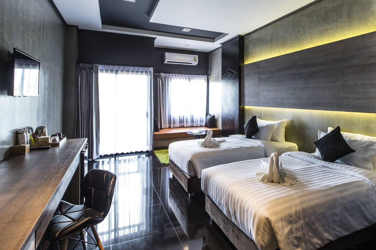 Quarto de hotel com escrivaninha, duas camas, área de estar e uma janela grande.
