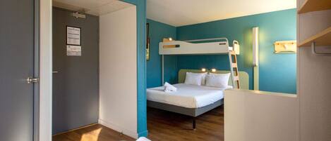 Quarto de hotel com beliche, uma cama de solteiro, porta, ventilação, luminária e piso de madeira.