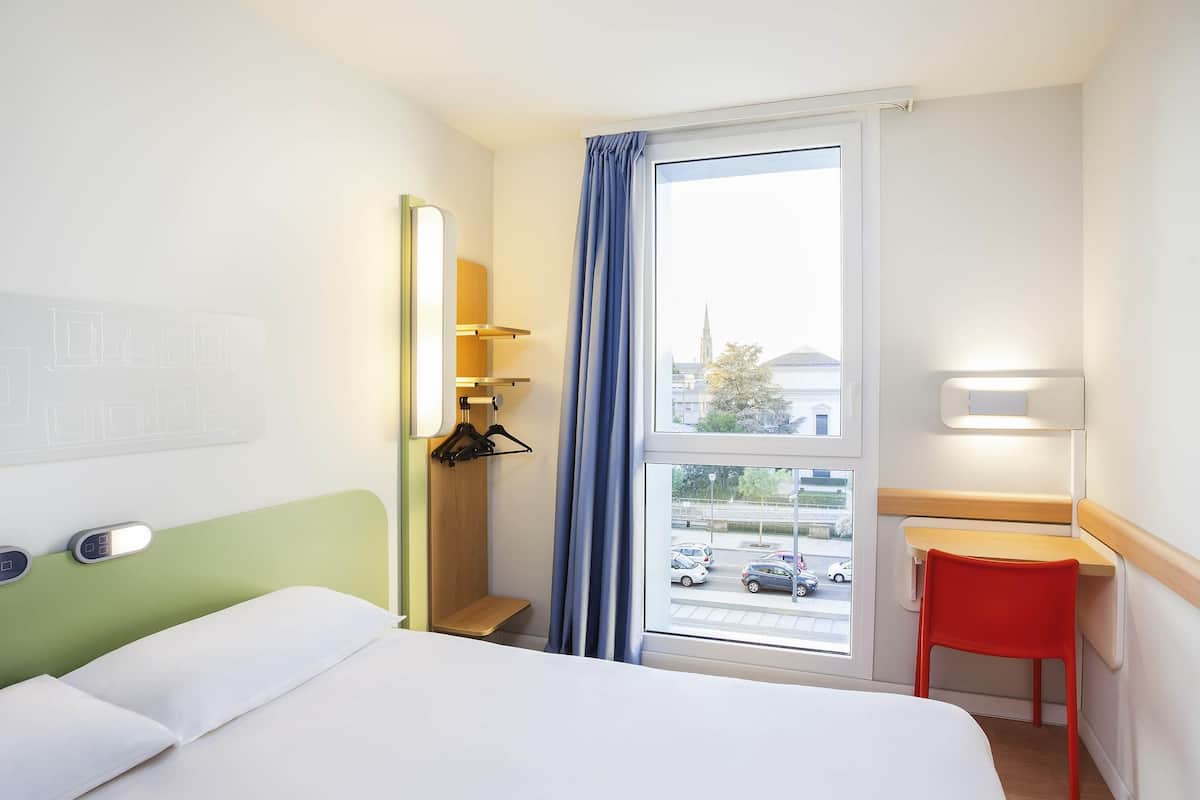 Quarto de hotel com cama, escrivaninha, cadeira, espelho, janela com vista para um edifício e carros, e um cabideiro.