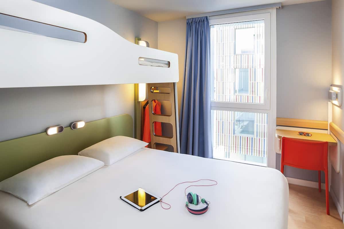 Quarto de hotel com beliche, escrivaninha, cadeira, janela com cortinas e mesa de cabeceira.