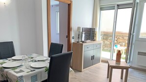 Uma área de refeições com uma mesa posta para quatro pessoas, um armário de madeira com uma televisão e vista para o exterior através de uma porta de vidro.