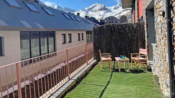 Uma varanda com vista para montanhas cobertas de neve, um muro de pedra e um pequeno espaço de estar com cadeiras e uma mesa.