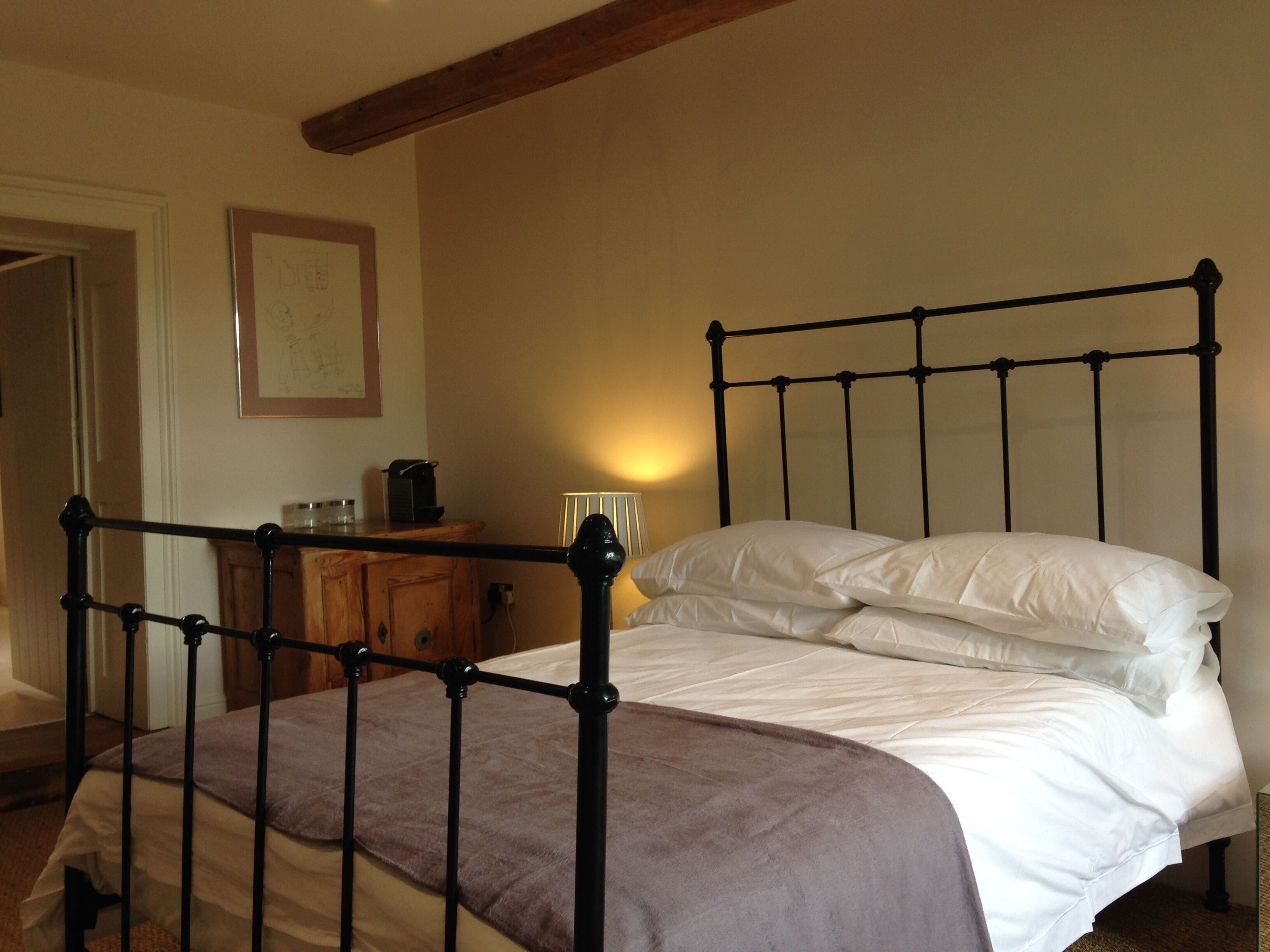 Uma cama com estrutura de metal e roupa de cama branca, uma mesa de cabeceira com abajur, e um armário de madeira em um quarto com porta e um quadro na parede.
