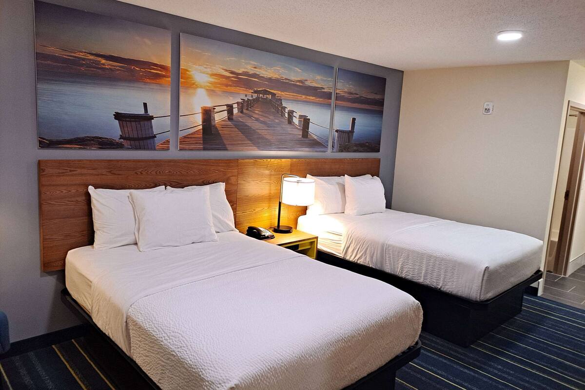 Quarto de hotel com duas camas, cabeceira de madeira, mesa de cabeceira com abajur e uma grande pintura com tema de pôr do sol na parede.