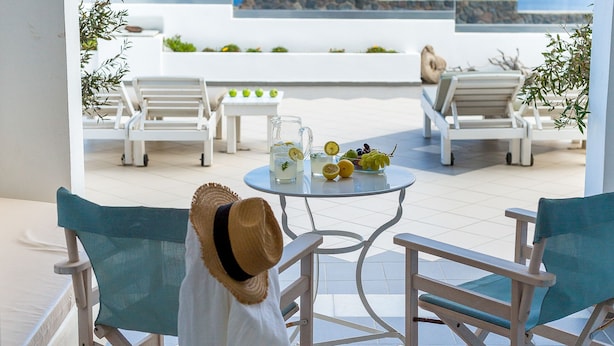 Um terraço com vista para o mar e montanhas, mobiliado com cadeiras brancas e uma mesa com frutas e uma bebida.