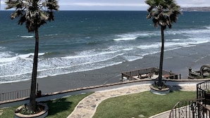 Vista para a praia com duas palmeiras, um caminho de pedra e um deck de madeira com bancos.