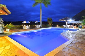 Piscina com borda de pedra, rodeada por cadeiras e guarda-sóis brancos, e uma palmeira ao fundo.