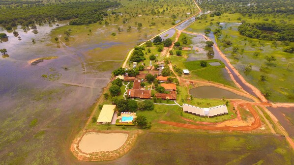 Vista aérea de uma área rural com um conjunto de edifícios, incluindo uma casa com piscina, cercada por campos e corpos d'água.