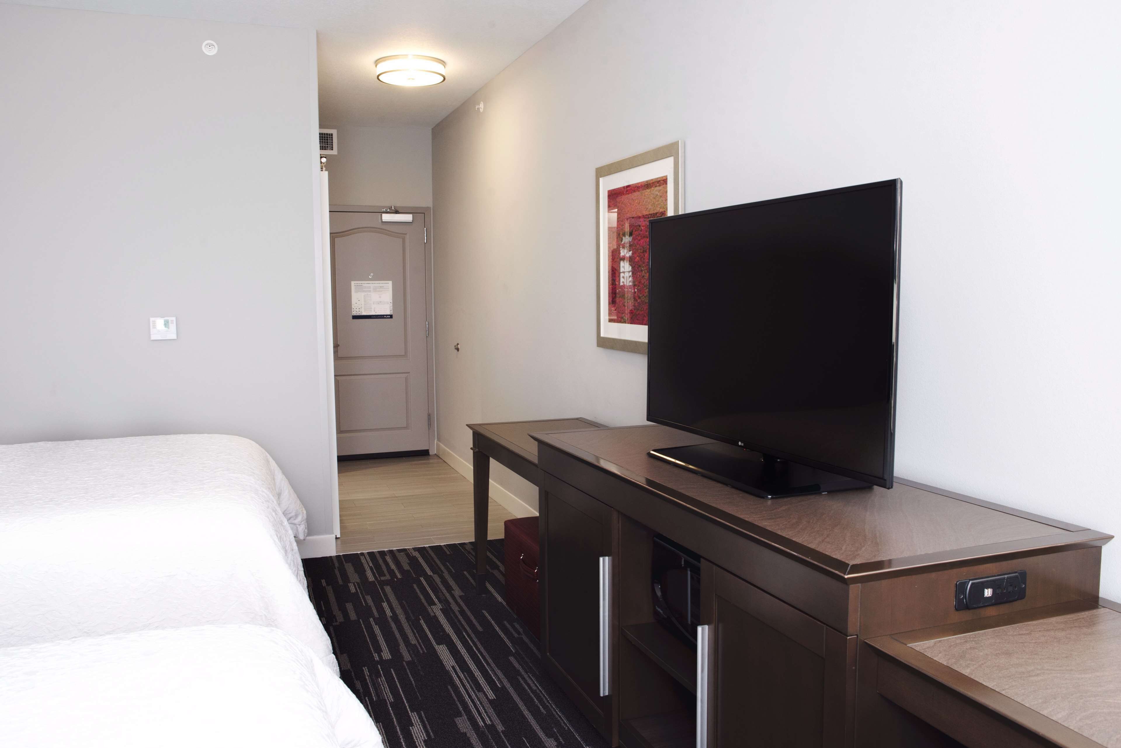 Quarto de hotel com uma cama, uma TV sobre uma cômoda de madeira, uma pequena mesa de escritório e uma porta que dá para outro quarto.