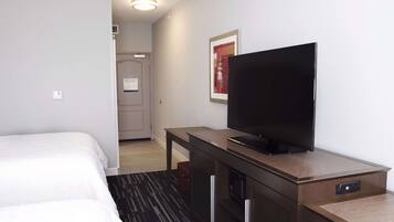 Quarto de hotel com uma cama, uma TV sobre uma cômoda de madeira, uma pequena mesa de escritório e uma porta que dá para outro quarto.
