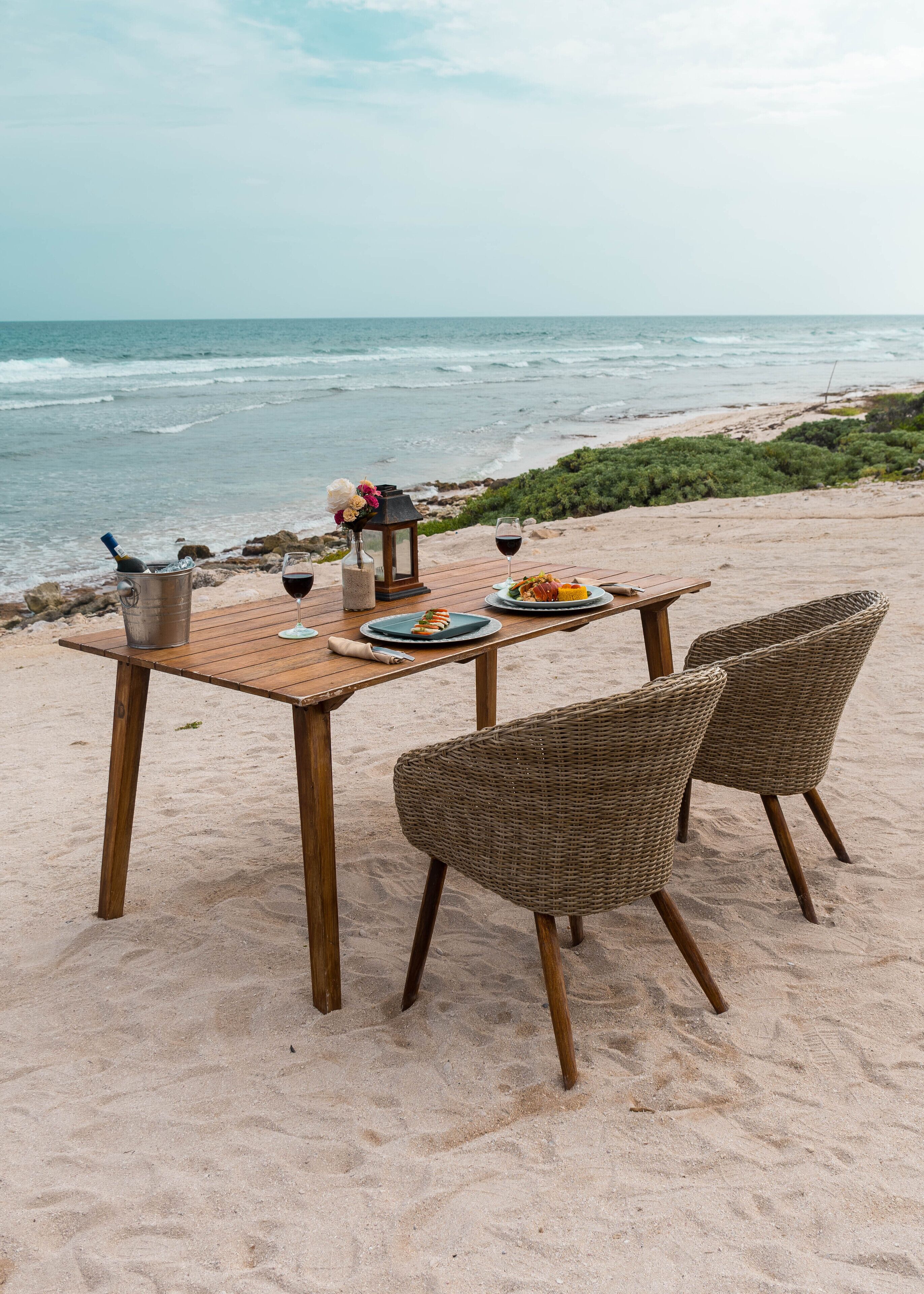Mesa de madeira montada em uma praia arenosa, com vista para o oceano. Há duas cadeiras de vime, uma garrafa de vinho, um copo de vinho e um prato de comida.