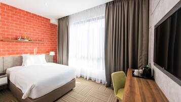 Quarto de hotel com uma cama grande, uma mesa com cadeira, uma televisão e uma janela com cortinas.