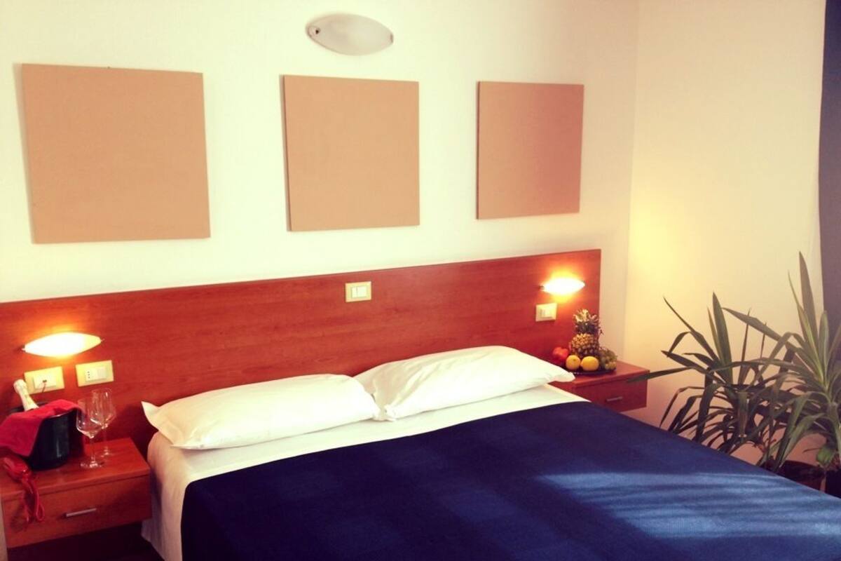 Quarto de hotel com cabeceira de madeira, cama com lençóis brancos, duas luminárias de cabeceira, telefone vermelho, uma taça de vinho e uma cesta de frutas.