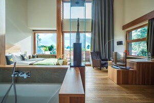 Quarto de hotel moderno com uma cama grande, área de estar com televisão, um banco de madeira e vista para o exterior.