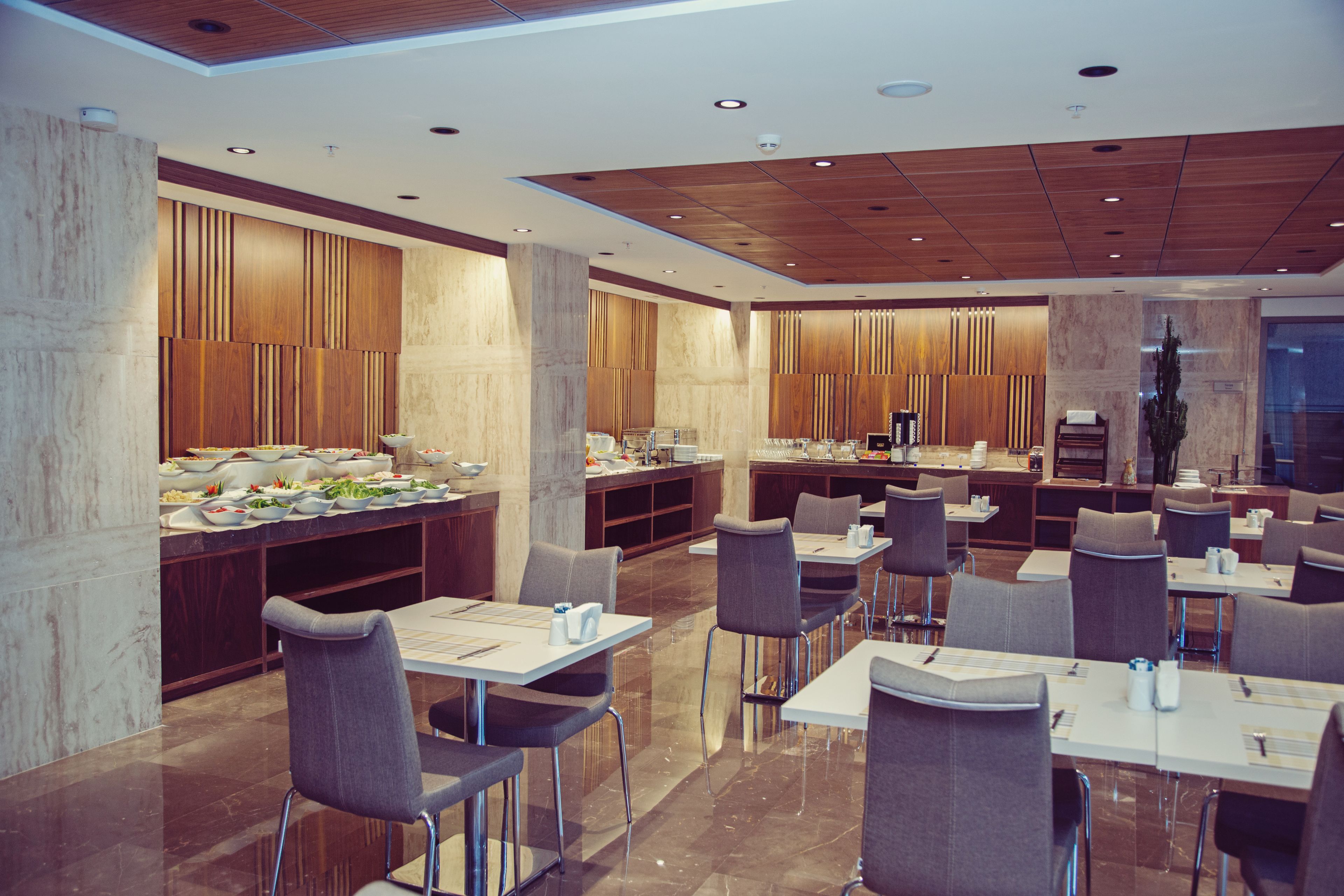 Uma área de refeições moderna com várias mesas e cadeiras, um buffet com diversos pratos e um detalhe no teto em madeira.