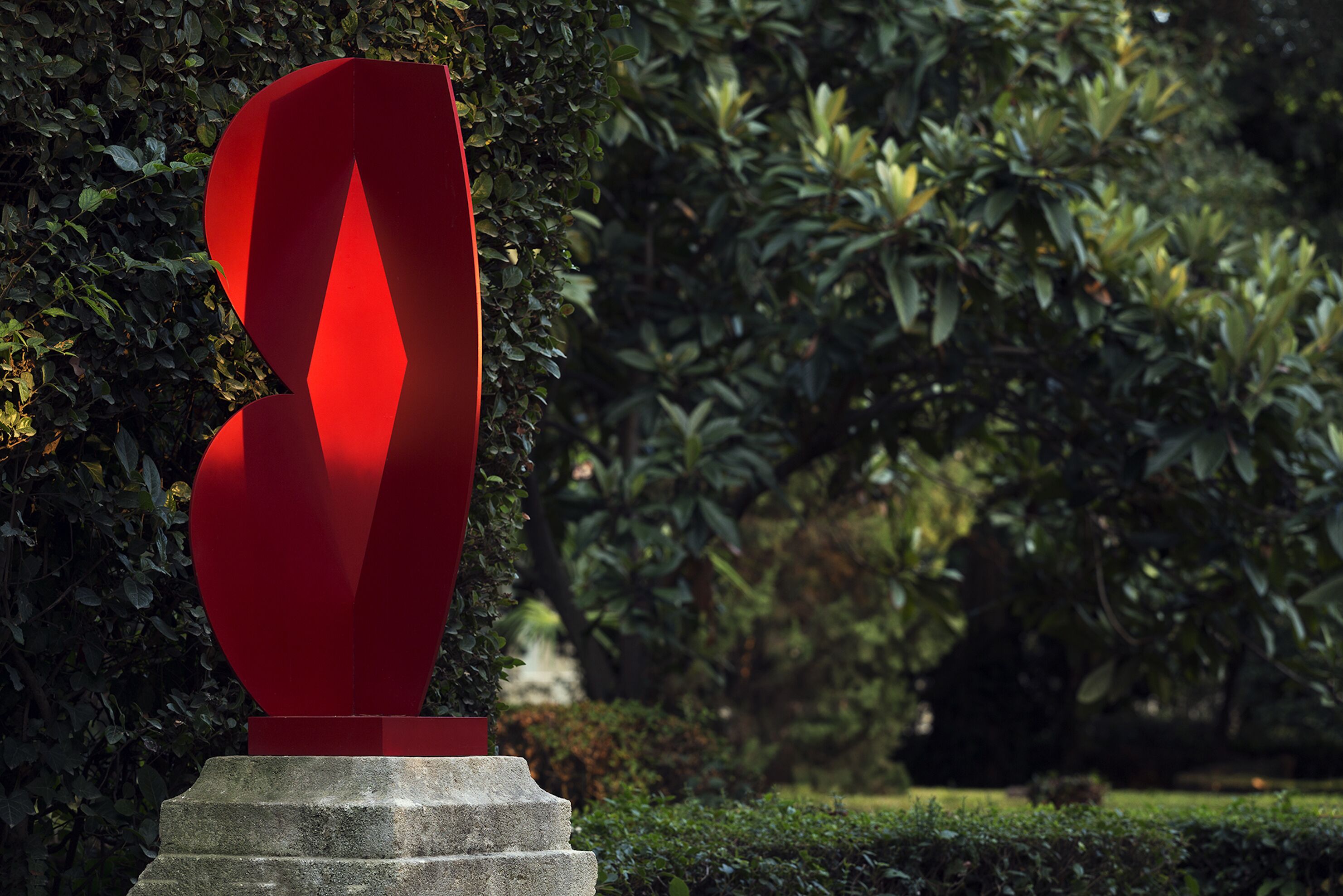 Uma escultura abstrata vermelha sobre um pedestal de pedra num jardim com folhagem densa ao fundo.