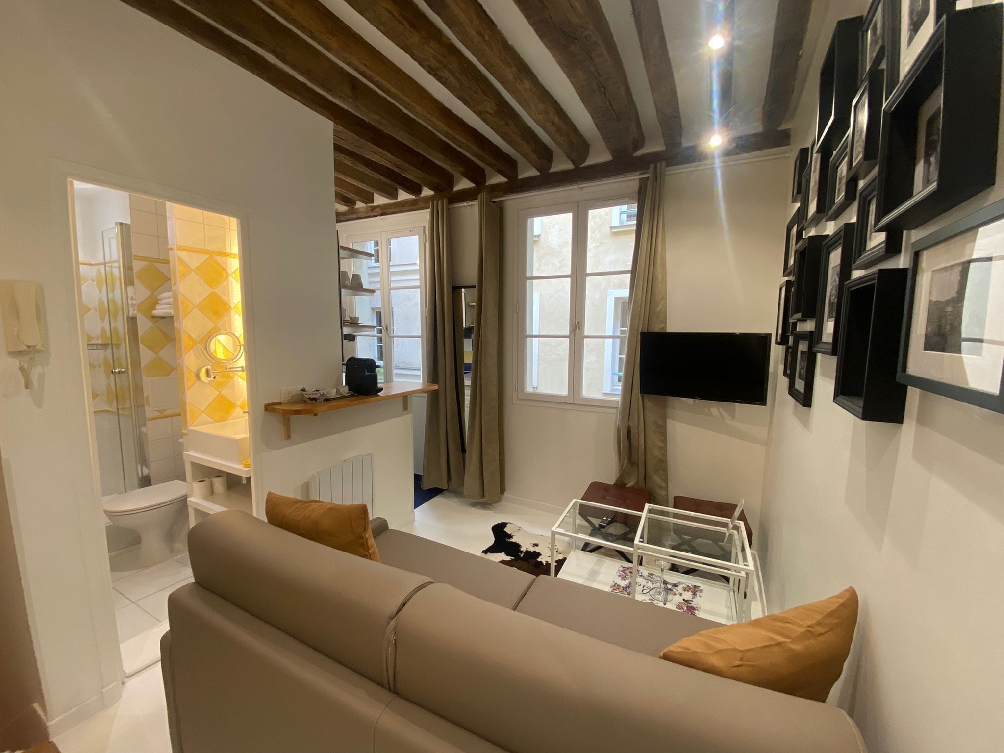 Sala de estar com sofá bege, mesa de centro e televisão fixada na parede. Há uma janela com cortinas, um radiador e uma estante com objetos.