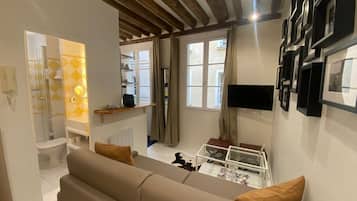 Sala de estar com sofá bege, mesa de centro e televisão fixada na parede. Há uma janela com cortinas, um radiador e uma estante com objetos.