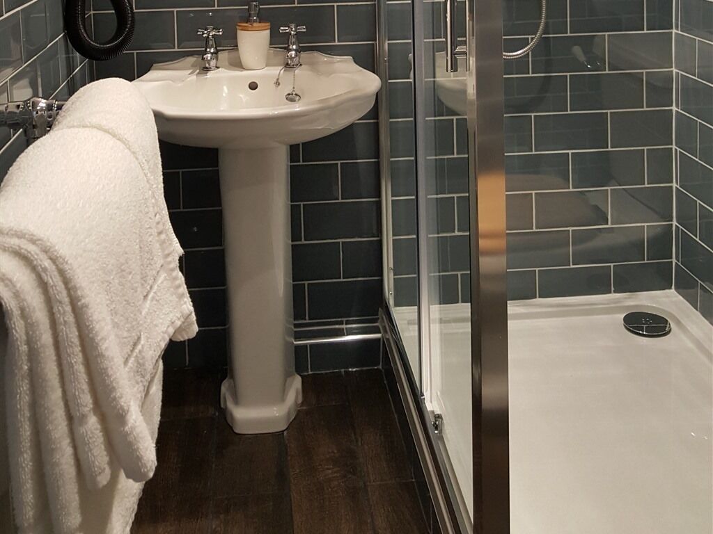 Casa de banho com lavatório pedestal branco, cabine de chuveiro em vidro e parede revestida a azulejos escuros.