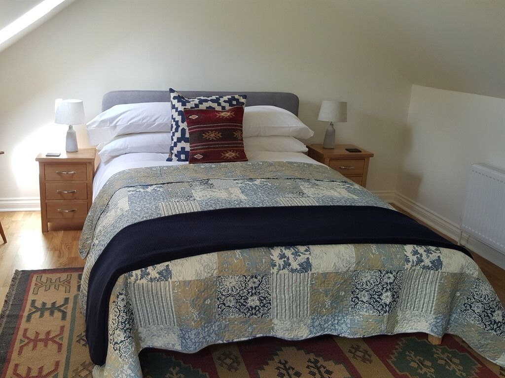 Cama bem arrumada com colcha estampada, duas luminárias de cabeceira, mesa de cabeceira de madeira e tapete estampado sobre piso de madeira.