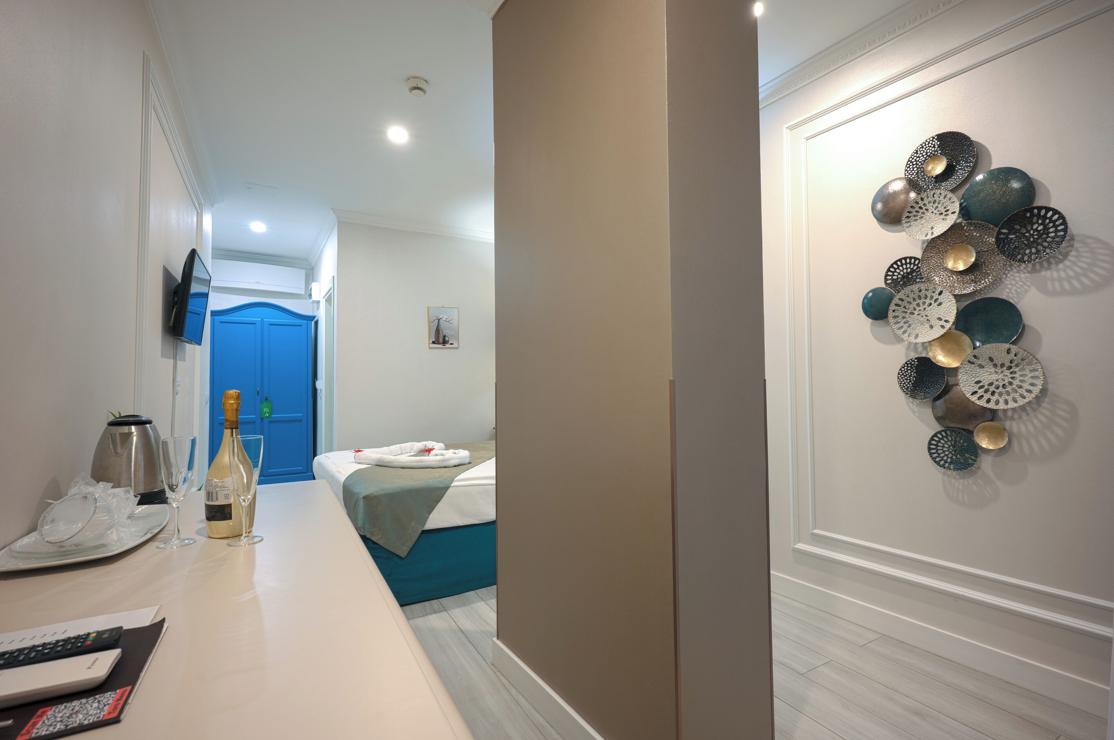 Quarto de hotel com cama, mesa de cabeceira com garrafa e copos, televisão na parede e um objeto decorativo na parede.