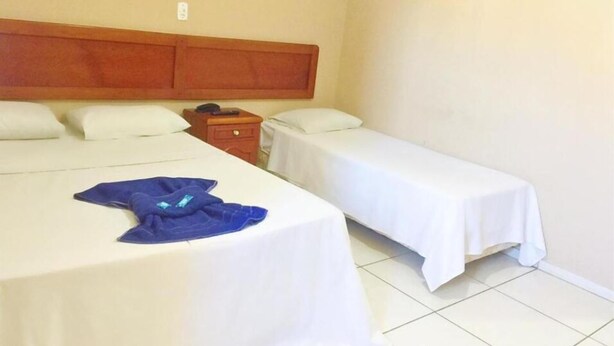 Quarto de hotel com duas camas, cabeceira de madeira, mesa de cabeceira e piso de azulejos brancos.