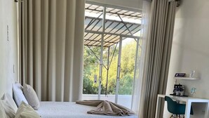 Quarto de hotel moderno com uma cama grande, cortinas bege, vista para vegetação e uma pequena mesa com cadeira.